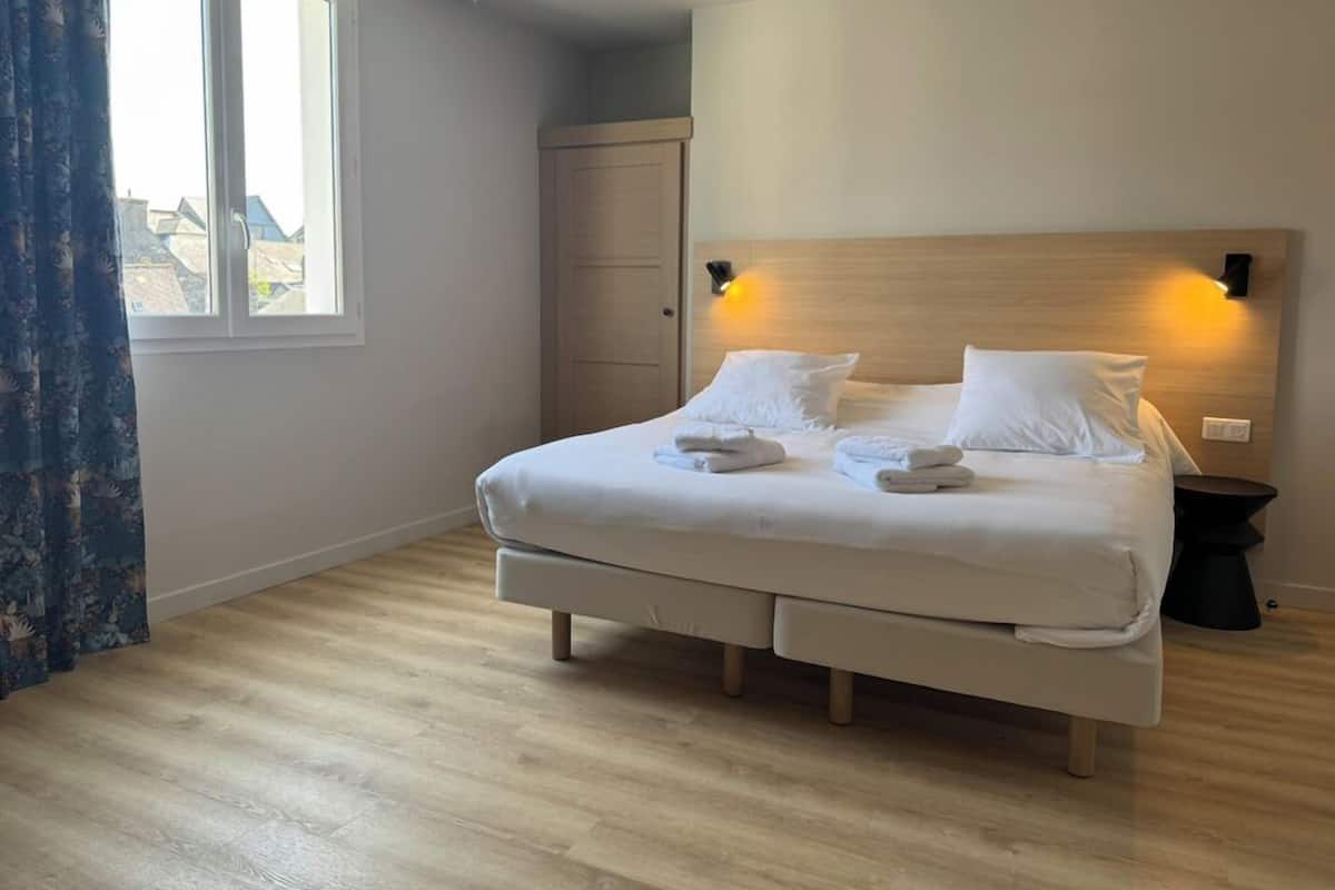Uma cama bem arrumada com almofadas e toalhas dobradas, uma mesa de cabeceira com abajur e uma janela com cortinas.