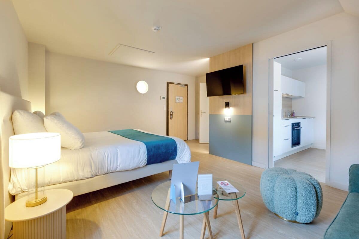 Quarto de hotel moderno com cama, mesa de cabeceira, abajur, mesa de centro de vidro, um banquinho e vista para a área da cozinha.