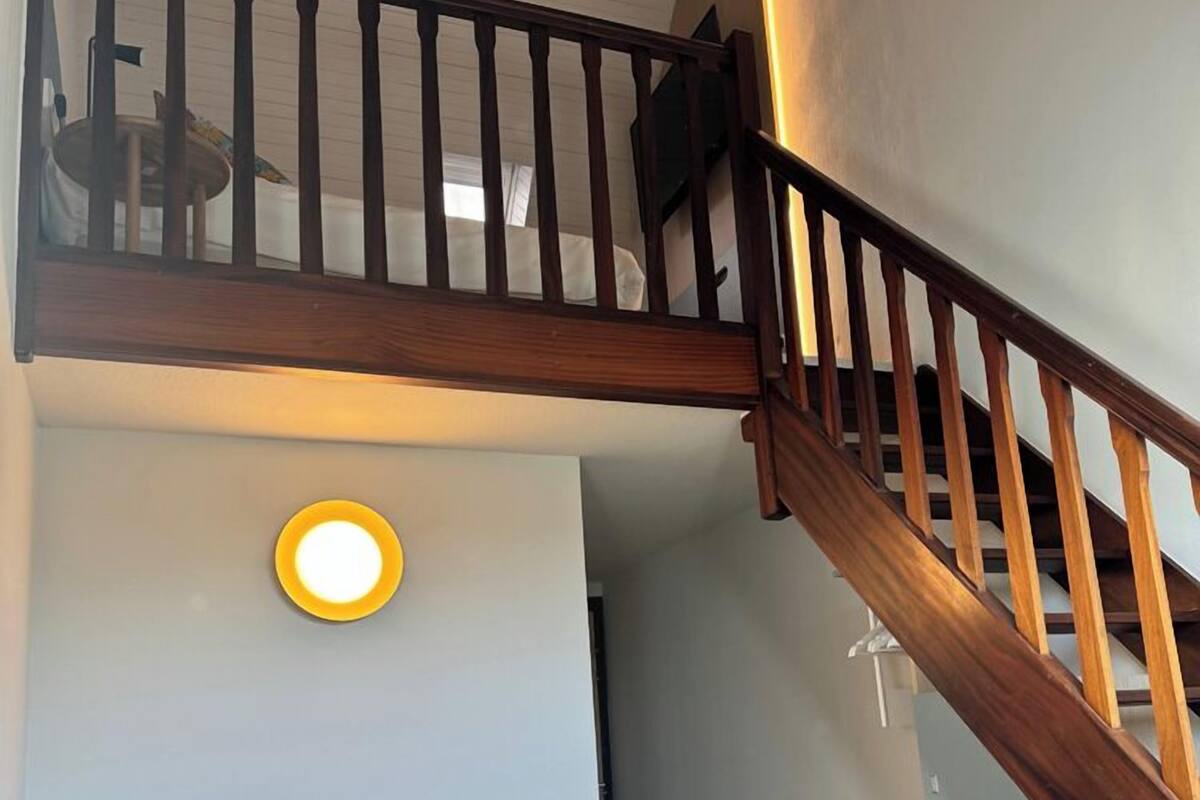 Um interior moderno com escada de madeira, sofá cinza e luminária embutida no teto.