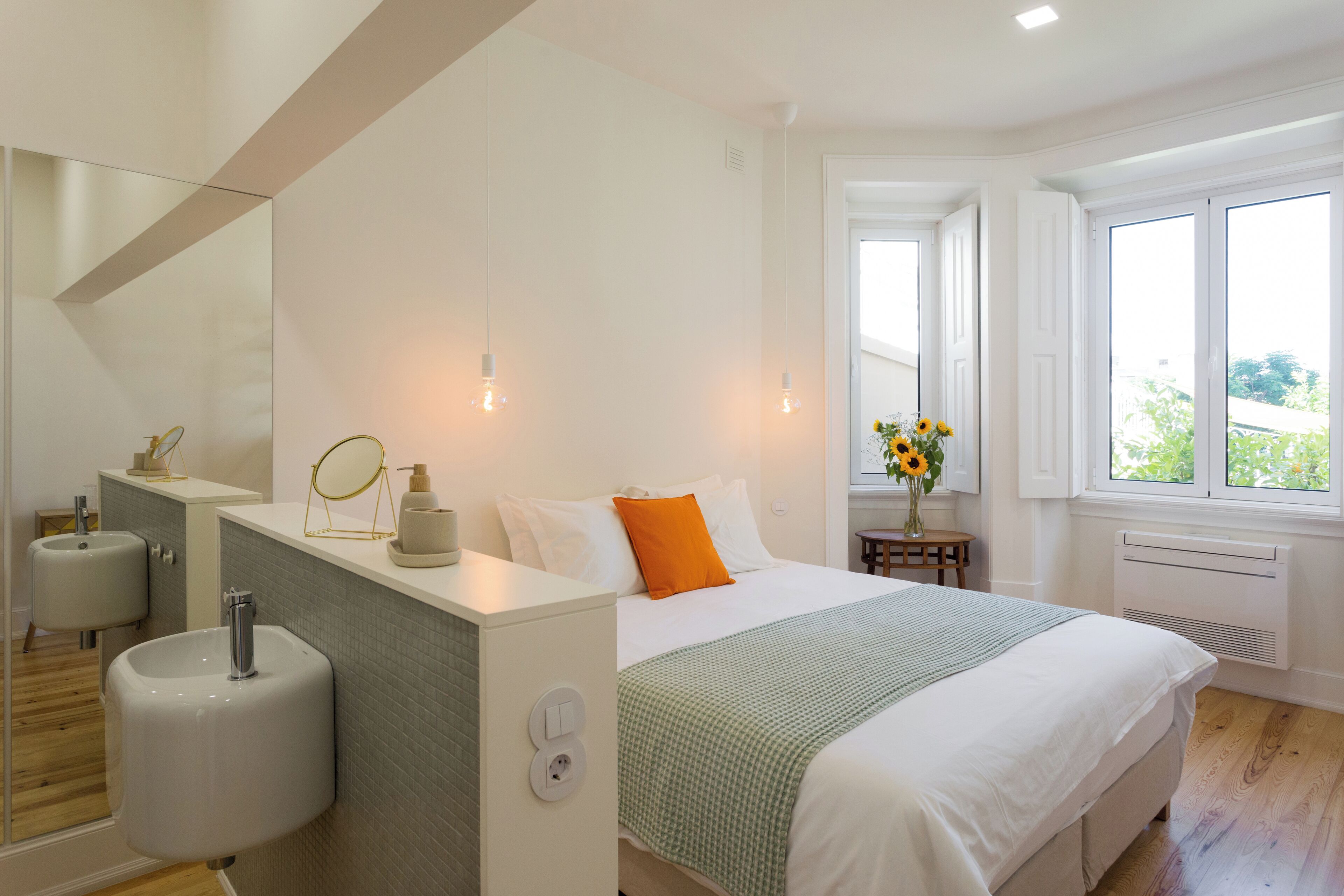 Quarto de hotel moderno com uma cama grande, uma mesa de cabeceira com um vaso de flores, banheiro com pia e espelho, e janelas amplas com vista para áreas verdes.