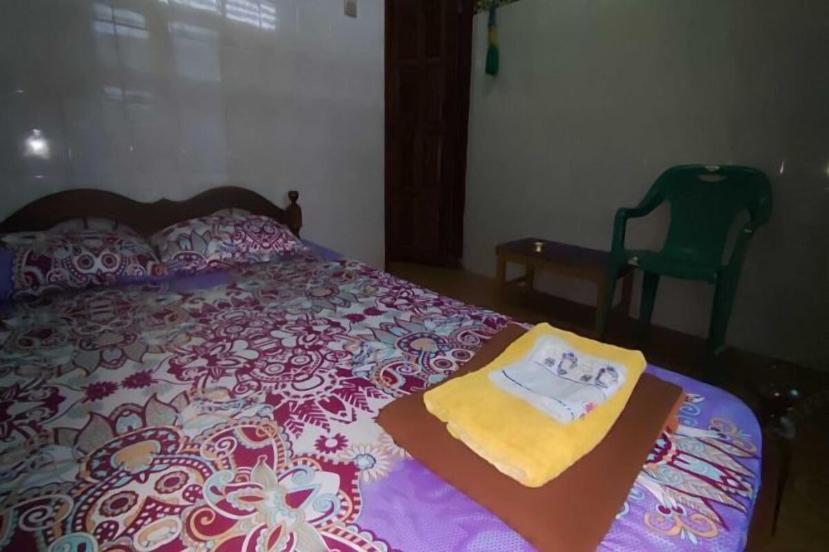 Quarto com cama coberta por um edredom estampado, cabeceira de madeira, uma cadeira de plástico verde e uma pequena mesa de madeira.
