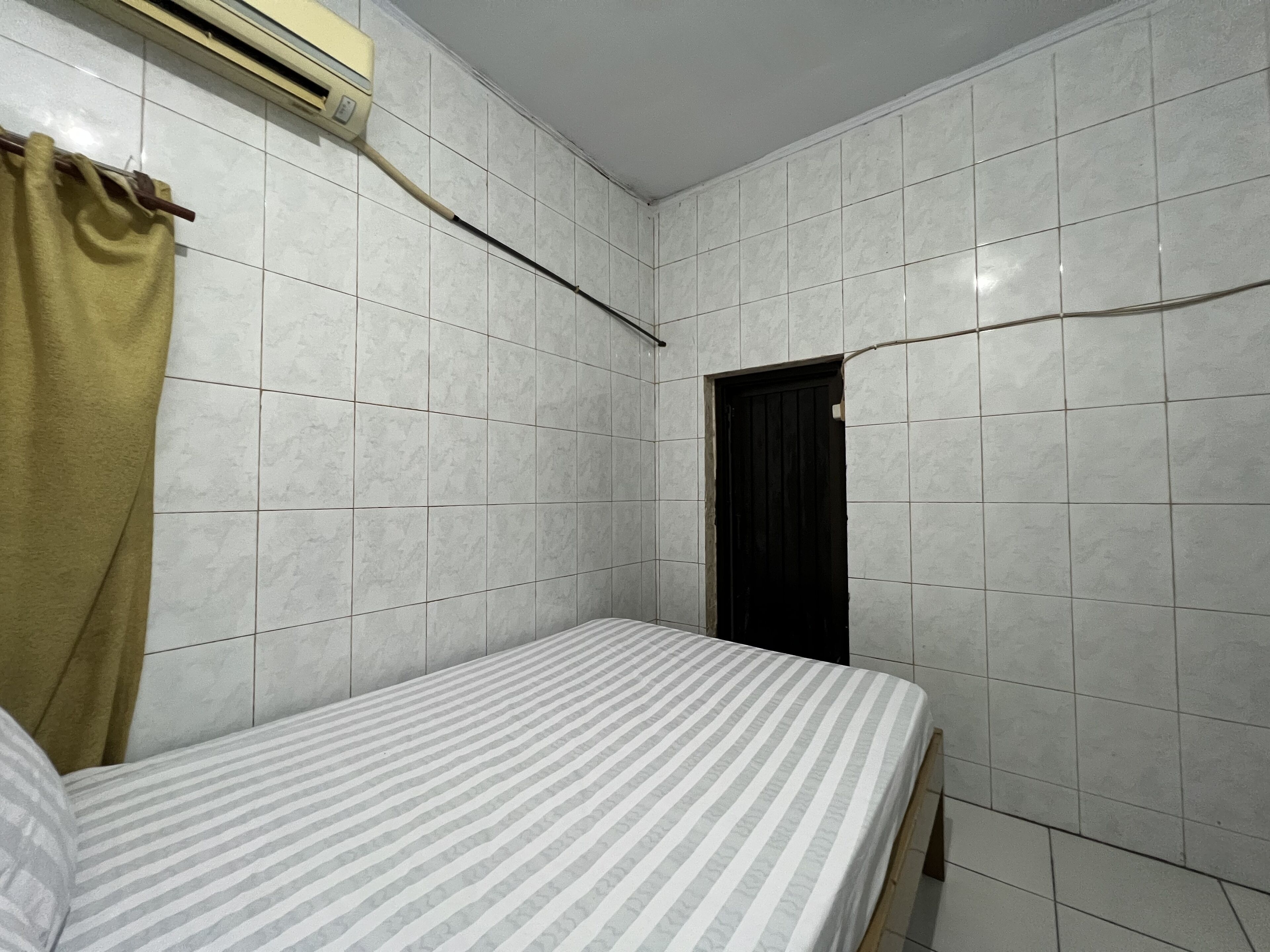 Um quarto pequeno, com piso de azulejos, uma cama de solteiro, uma cortina e uma porta.