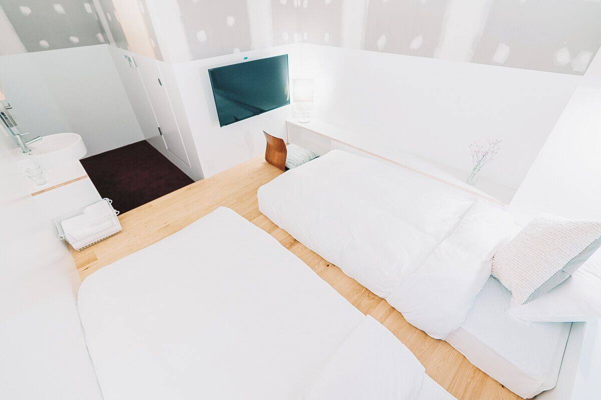 Quarto de hotel moderno com uma cama grande, uma mesa de cabeceira, televisão na parede e banheiro com pia e espelho.