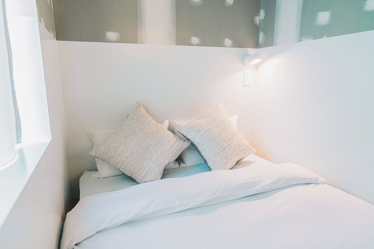 Cama com roupa de cama branca e duas almofadas, janela com persianas e luminária fixada na parede.