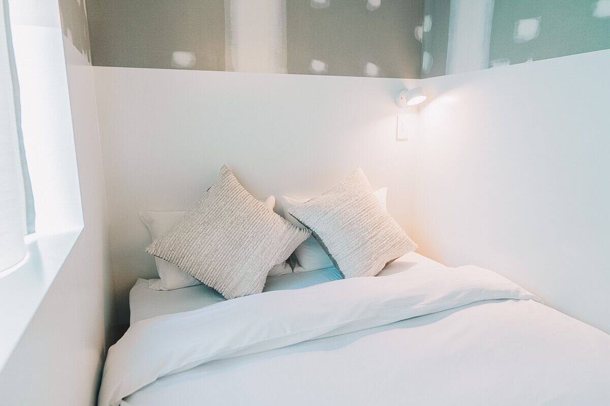 Cama bem arrumada com lençóis brancos e duas almofadas, janela com persianas e luminária fixada na parede.