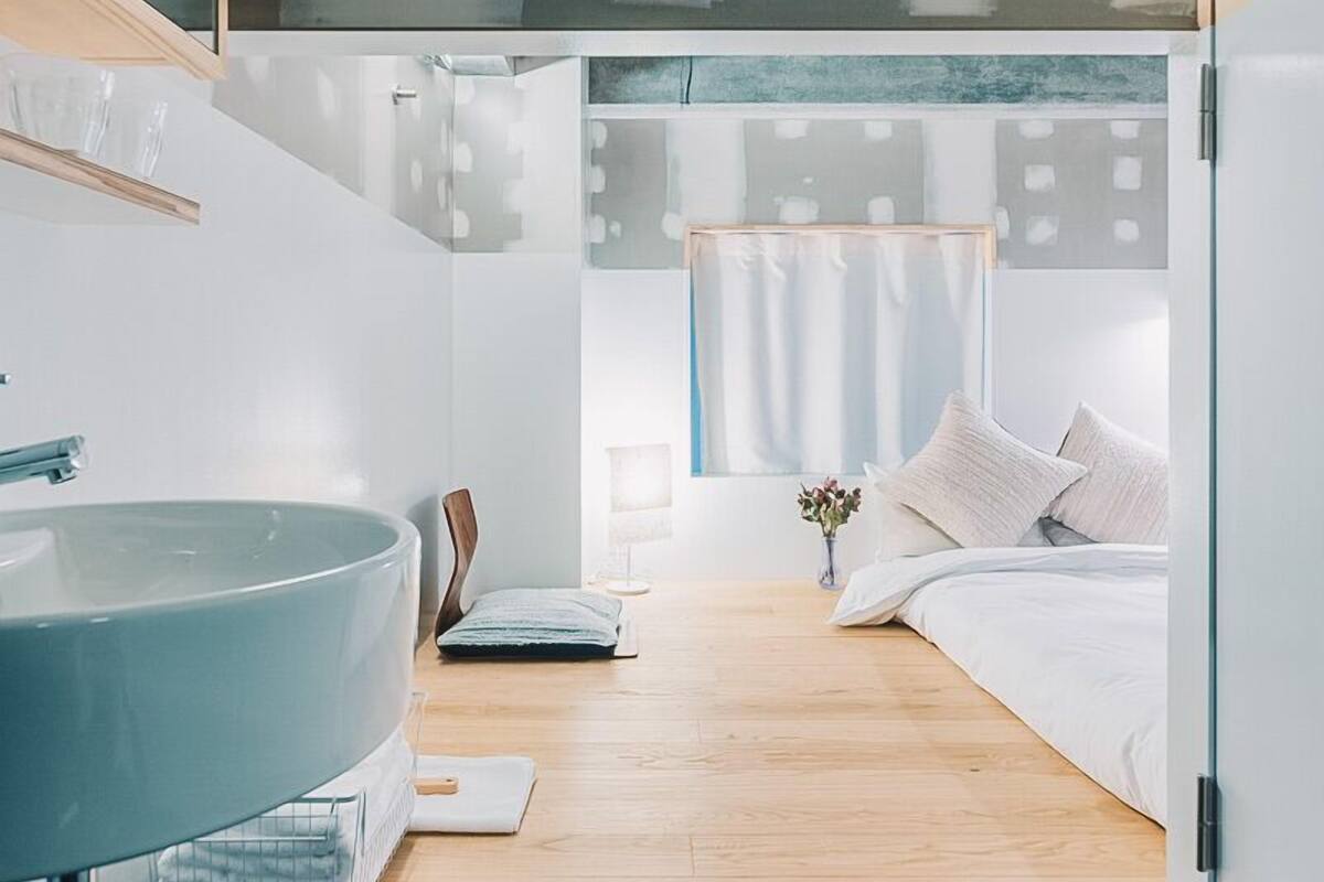 Quarto compacto com uma janela grande, mesa de cabeceira, cama e pia embutida.