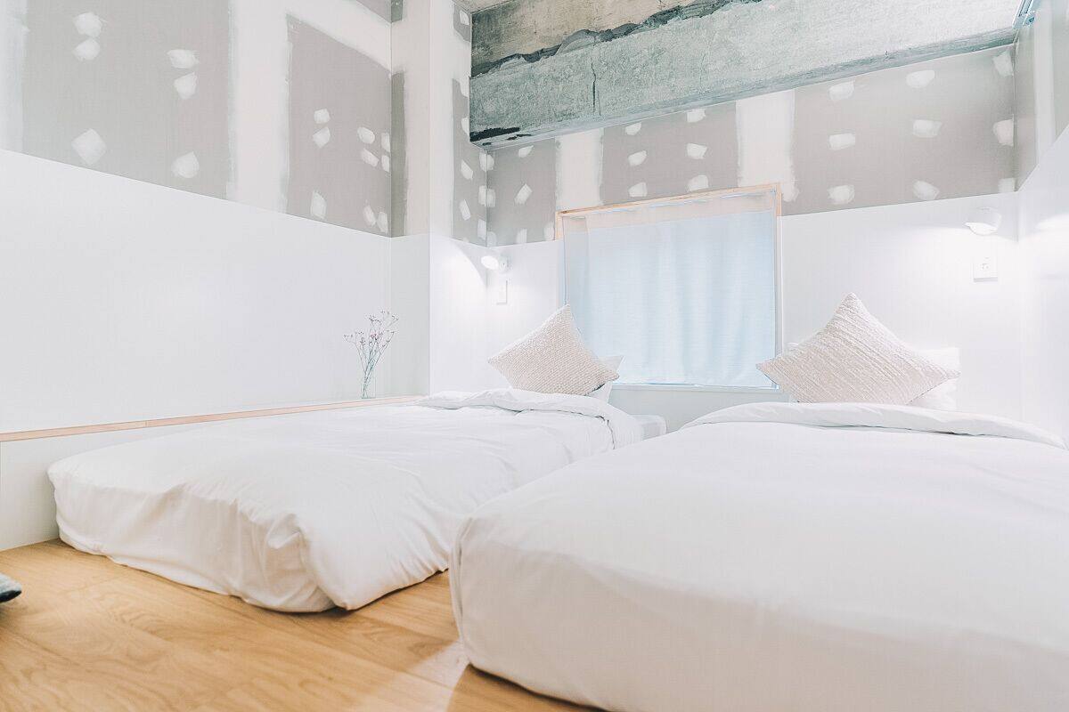 Quarto com duas camas, piso de madeira, janela com persianas e parede com textura.