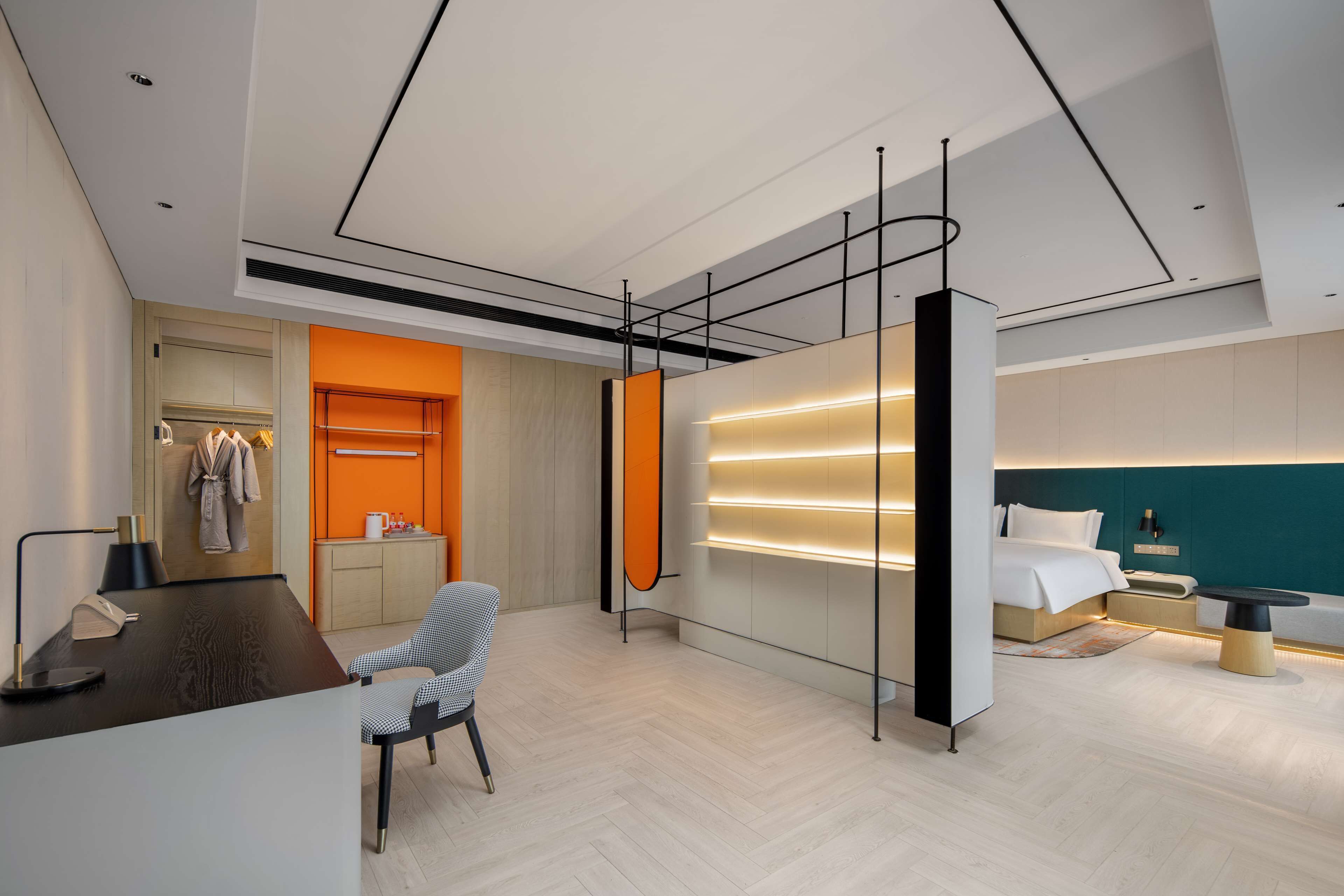 Quarto de hotel moderno com piso de madeira, uma escrivaninha com luminária, uma cadeira, uma cama com cabeceira, mesa de cabeceira e prateleira na parede.