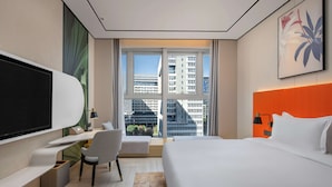 Quarto de hotel moderno com uma cama grande, uma escrivaninha, uma cadeira, uma TV e uma janela com vista para a cidade.