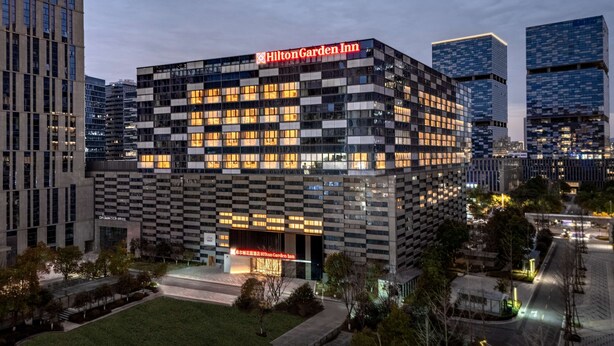 Um hotel moderno com fachada xadrez, janelas iluminadas e um sinal proeminente de Hilton Garden Inn.