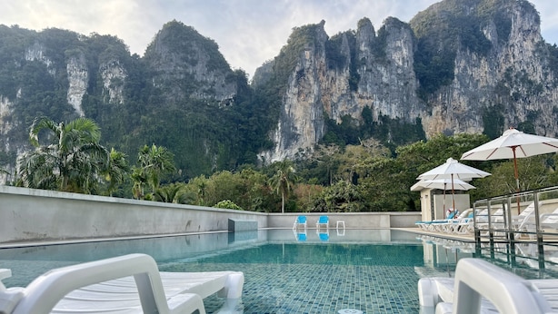 Piscina com espreguiçadeiras e guarda-sóis, cercada por um muro de concreto, com vista para falésias rochosas e vegetação.