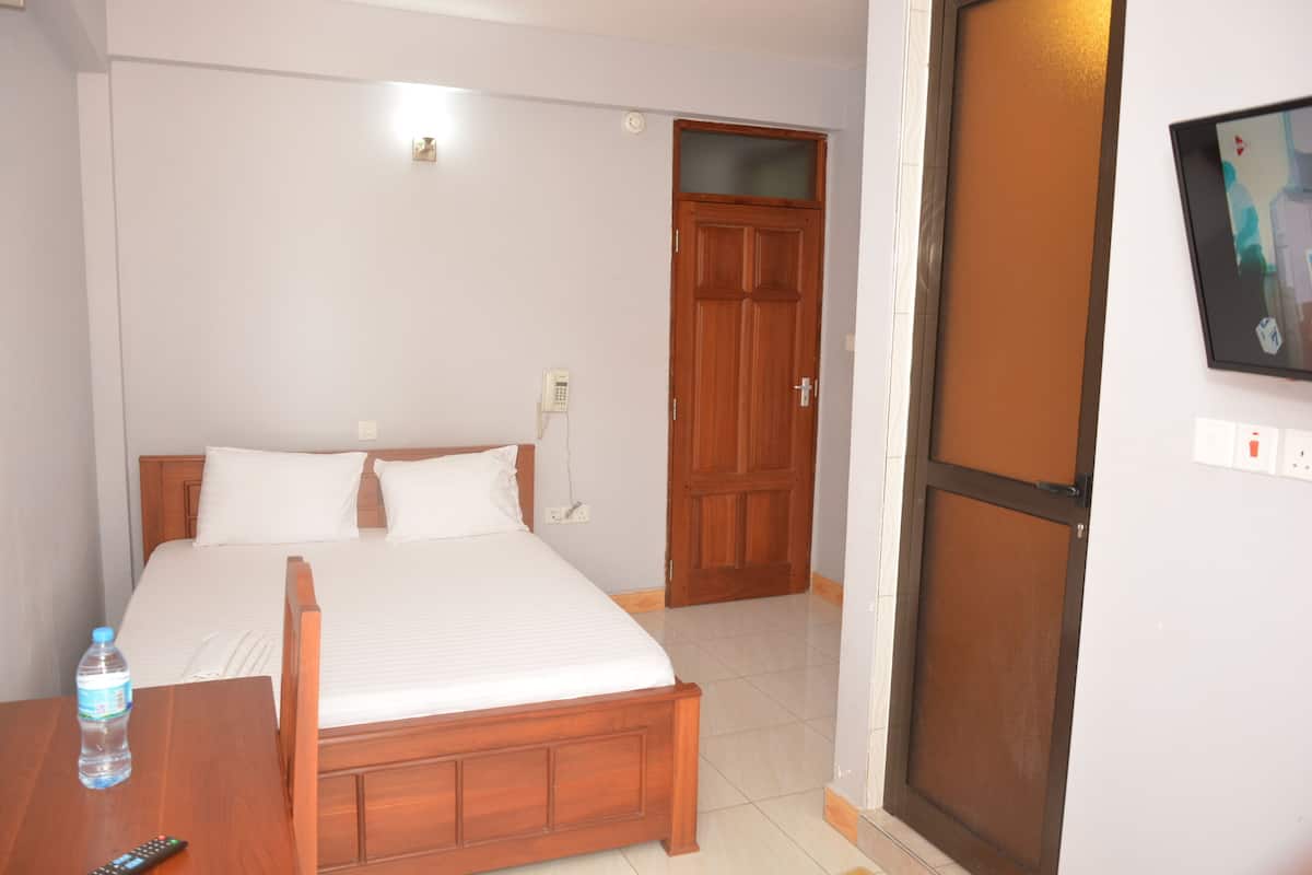 Quarto com cama de madeira, mesa de cabeceira com uma garrafa e controle remoto, telefone fixado na parede e porta com painel de vidro.