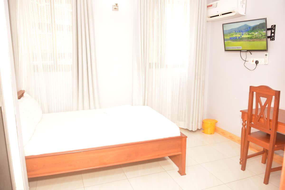 Quarto com cama e cadeira de madeira, televisão na parede e janela com cortinas leves.
