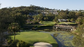 Um campo de golfe com um lago, uma ponte de madeira e um edifício ao fundo.