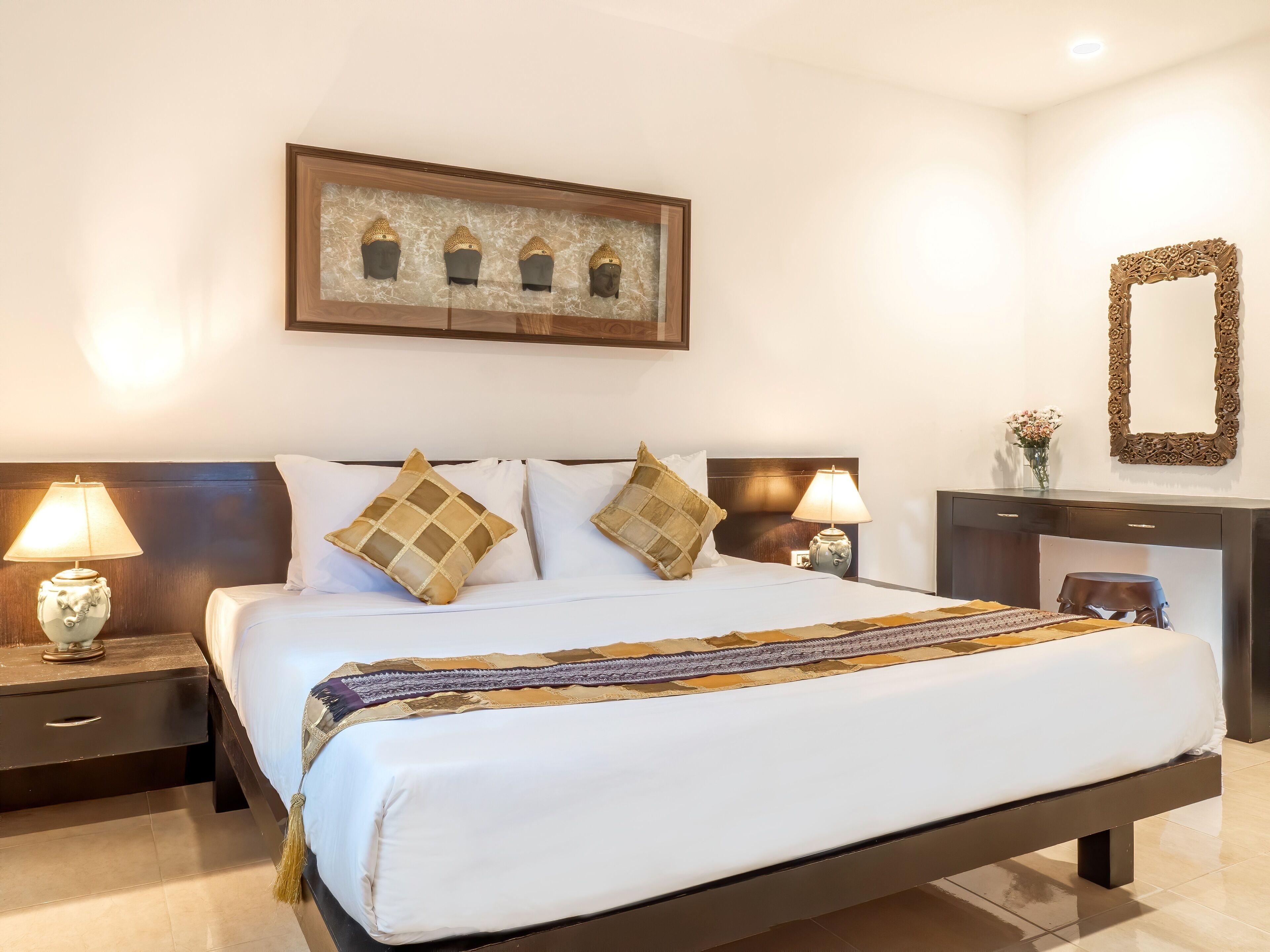 Cama bem arrumada com edredom branco e almofadas estampadas, cabeceira de madeira, mesa de cabeceira com abajur, cômoda com gavetas e um quadro emoldurado sobre a cama.