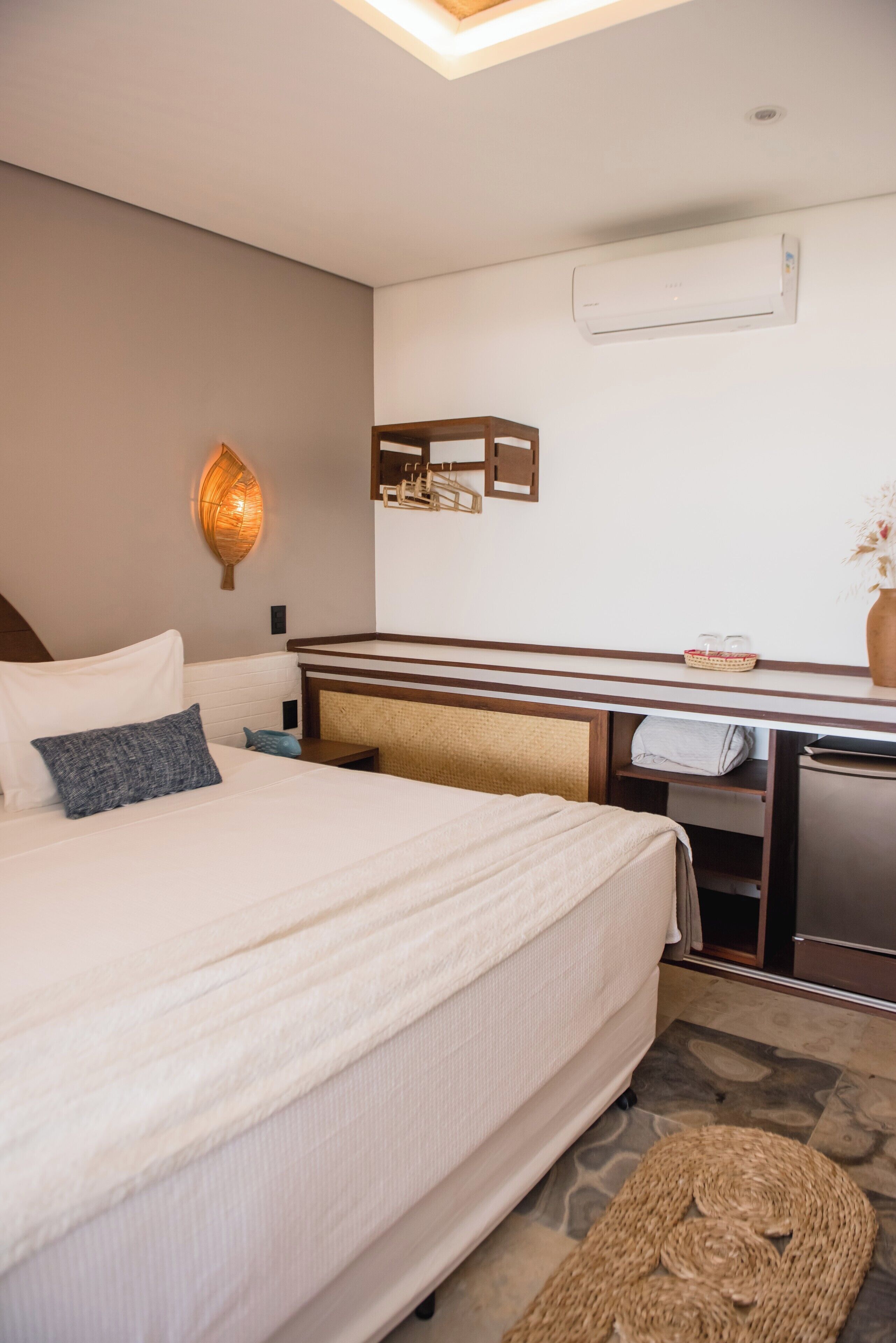 Quarto de hotel com uma cama grande, uma mesinha de cabeceira, ar-condicionado fixado na parede e uma luminária decorativa.