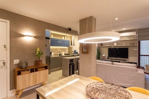 Cozinha moderna com ilha central, área de jantar com mesa de madeira e sala de estar com TV e assentos.