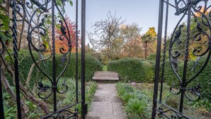 Caminho no jardim que leva a um banco cercado por grades de metal elaboradas e vegetação exuberante.