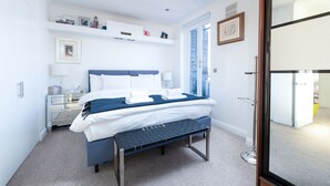 Quarto moderno com cama, mesa de cabeceira, banqueta e porta de vidro que dá para outro cômodo.