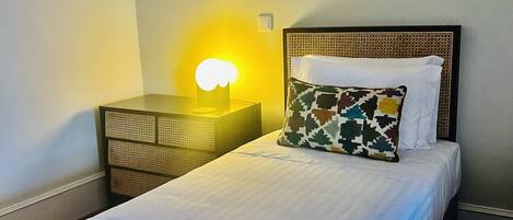 Cama bem arrumada com edredom branco e almofada estampada, luminária de cabeceira e uma pequena mesa de cabeceira de madeira com uma luminária.