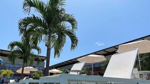 Área de piscina com espreguiçadeiras e guarda-sóis, cercada por palmeiras e edifícios modernos.