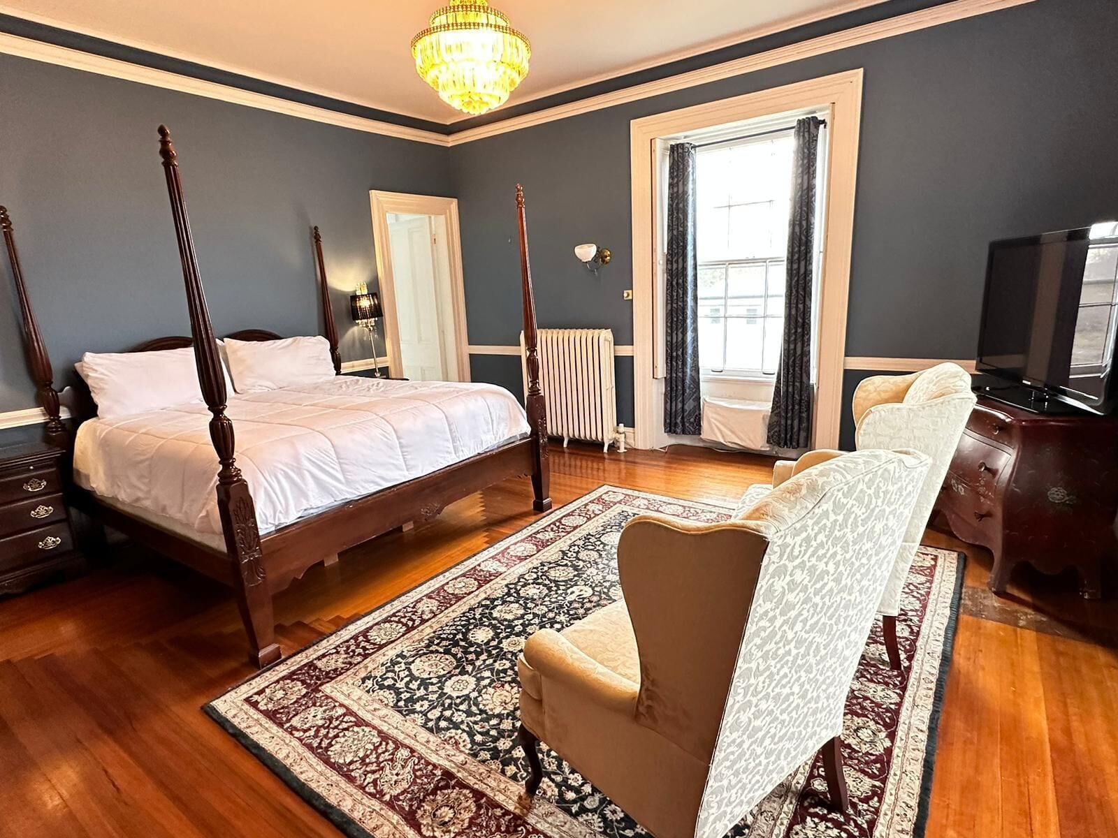 Quarto com cama de dossel, lustre, televisão e uma grande janela com cortinas.