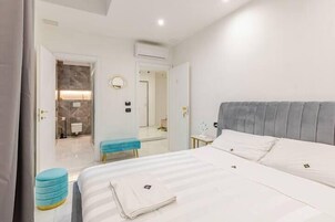 Quarto de hotel com uma cama grande, uma mesa de cabeceira, um espelho e um banheiro visível por uma porta aberta.