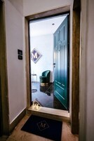 Entrada de um hotel com uma porta verde-água, um tapete preto com um monograma e uma placa na parede com a letra 'M' em tamanho grande.
