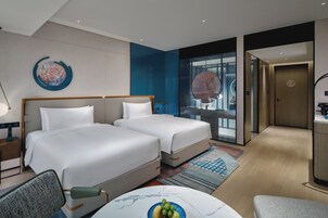 Quarto de hotel com duas camas, uma escrivaninha, uma cadeira, uma mesa redonda com uma tigela de frutas, uma grande pintura abstrata e vista da cidade através de uma parede de vidro.