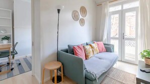 Sala de estar com sofá, mesa pequena e luminária de chão. Porta de vidro deslizante dá acesso a uma área externa.