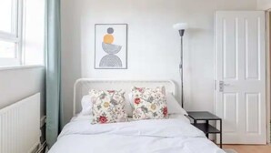 Quarto com cama, almofadas florais, mesa de cabeceira, abajur, porta e uma obra de arte na parede.