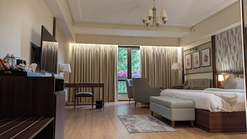 Quarto de hotel com cama, escrivaninha, cadeira, televisão e uma janela grande com cortinas.