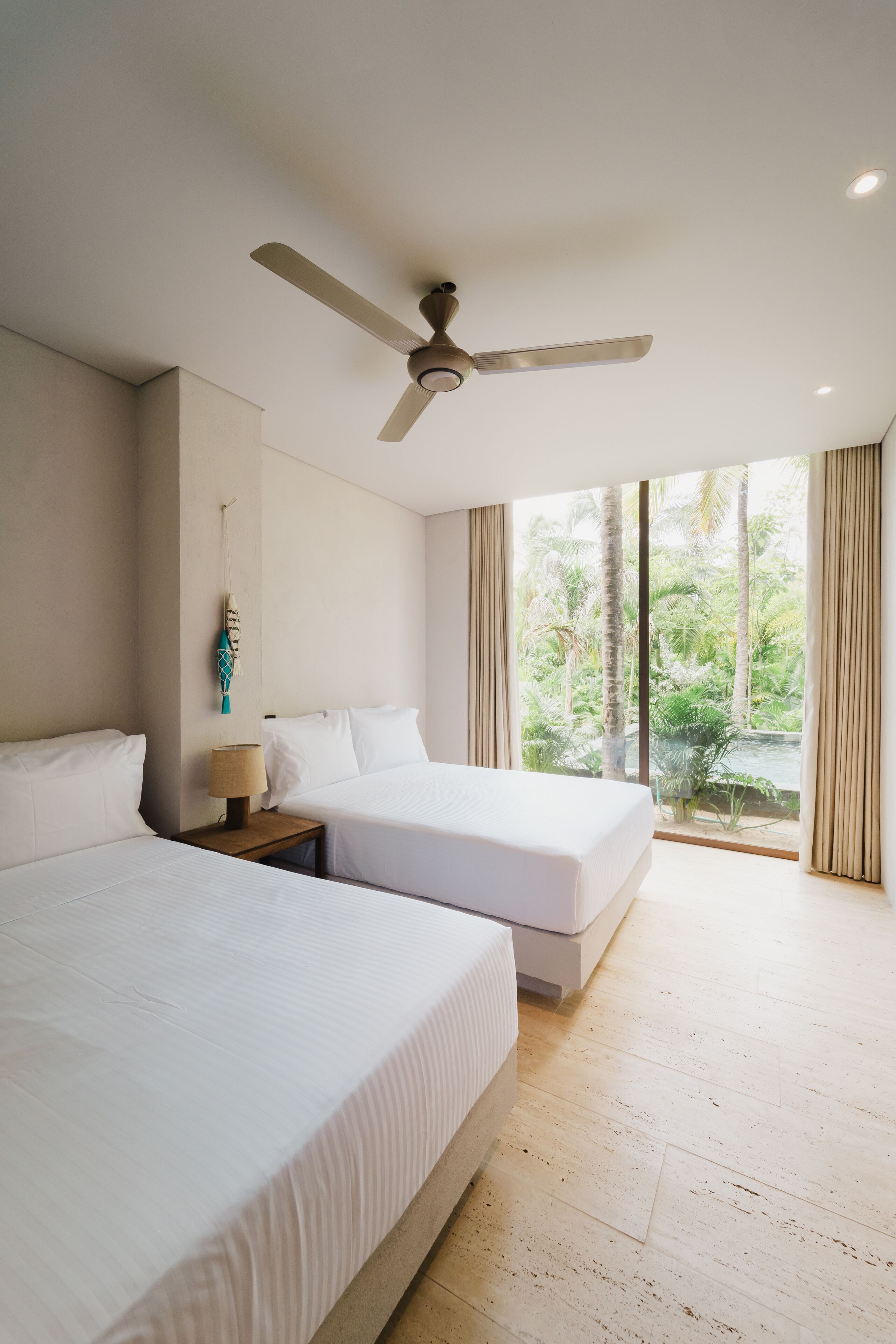 Quarto de hotel com duas camas, ventoinha no teto e uma grande janela com vista para o verde.