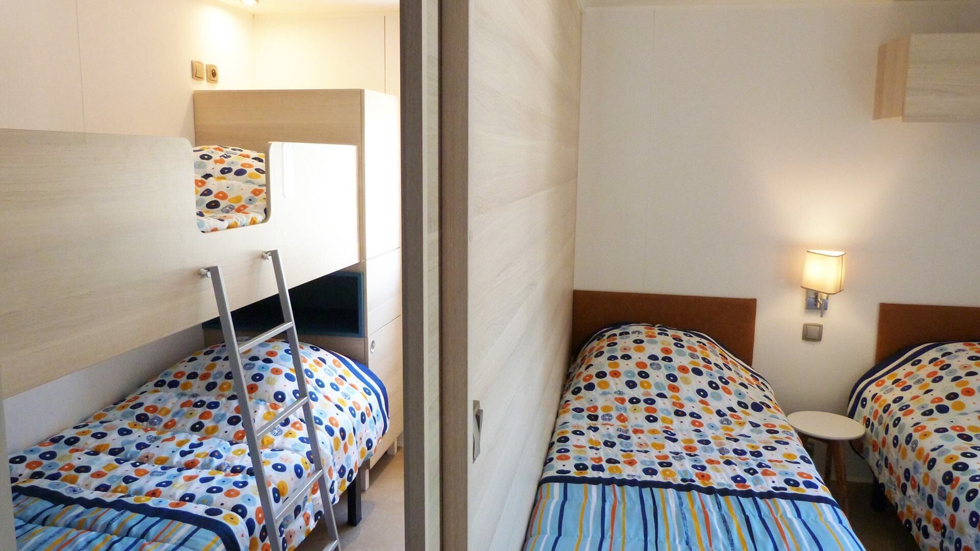 Quarto de dormitório com beliches e uma cama simples, ambos com roupa de cama em bolinhas. Há uma pequena mesa e uma luminária na parede.