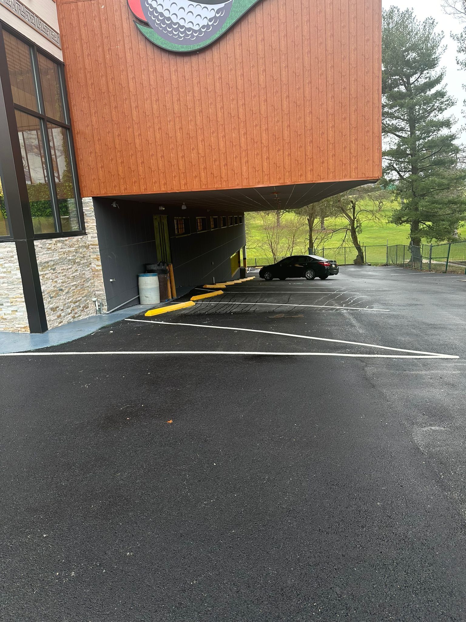 Um estacionamento com um carro preto estacionado ao longe, um edifício com fachada de madeira e um campo verde ao fundo.