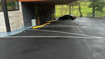 Um estacionamento com um carro preto estacionado ao longe, um edifício com fachada de madeira e um campo verde ao fundo.