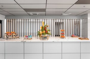 Uma área de recepção com balcão branco, persianas verticais e diversos objetos, como um vaso de flores, uma pequena prateleira com lanches e uma escada com mais lanches.