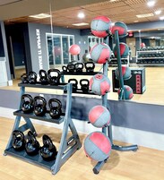 Uma academia com kettlebells e bolas medicinais em suportes, um espelho refletindo os equipamentos e uma placa com texto na parede.