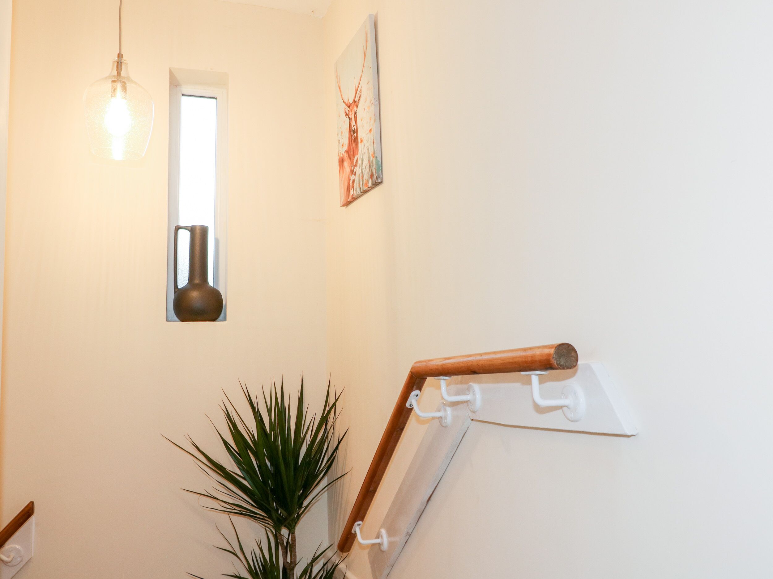 Uma escadaria com corrimão, um vaso de planta, um luminário fixado na parede e uma pequena janela.