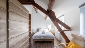 Quarto com parede de ripas de madeira, cama com roupa de cama branca, cadeira de madeira com travesseiro amarelo e teto inclinado com vigas aparentes.