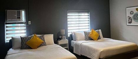 Quarto de hotel com duas camas, uma pequena mesa de cabeceira com abajur e um ar condicionado fixado na parede.