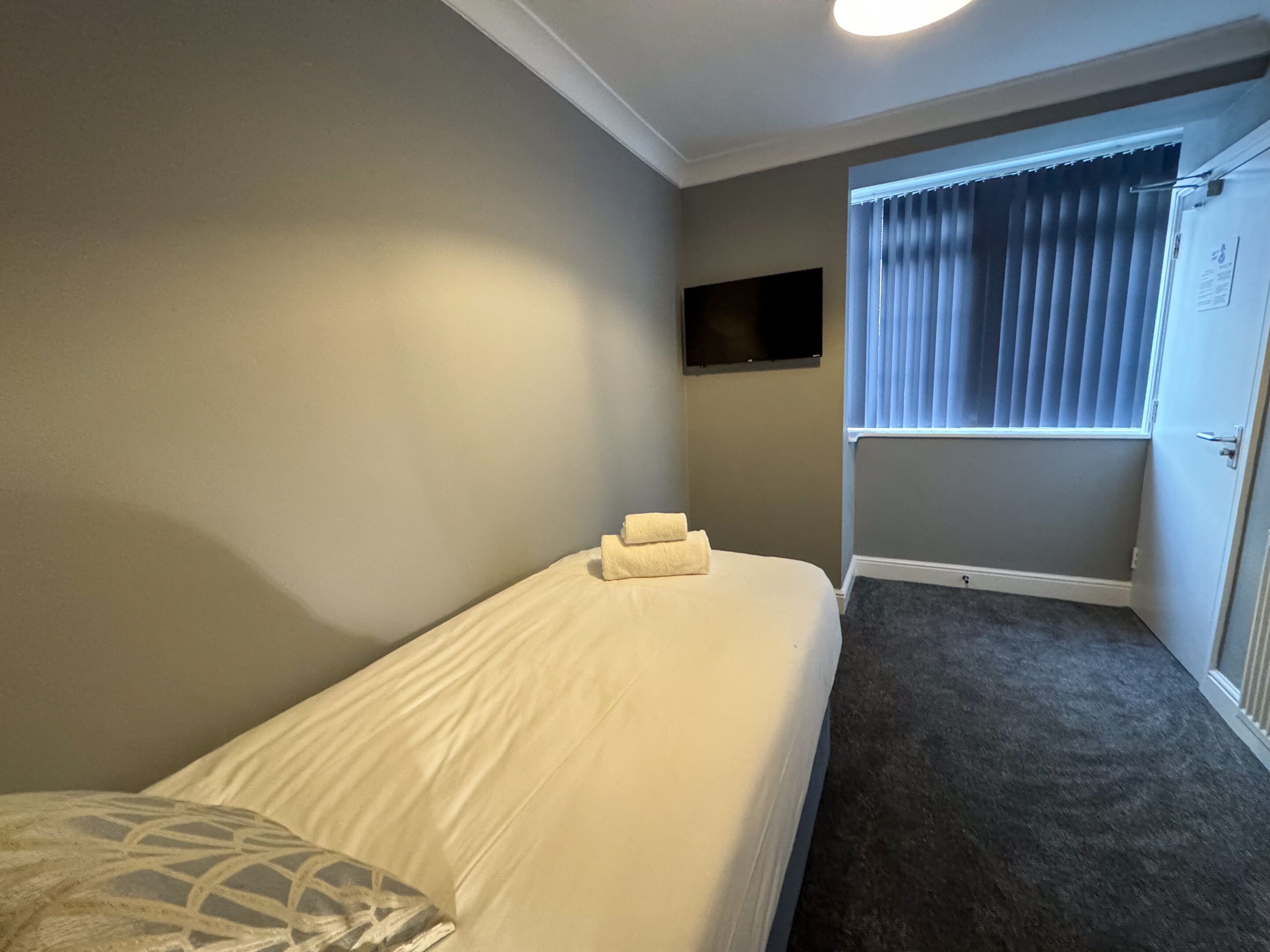 Quarto de hotel com uma cama de solteiro, televisão de tela plana na parede e janela com persianas.