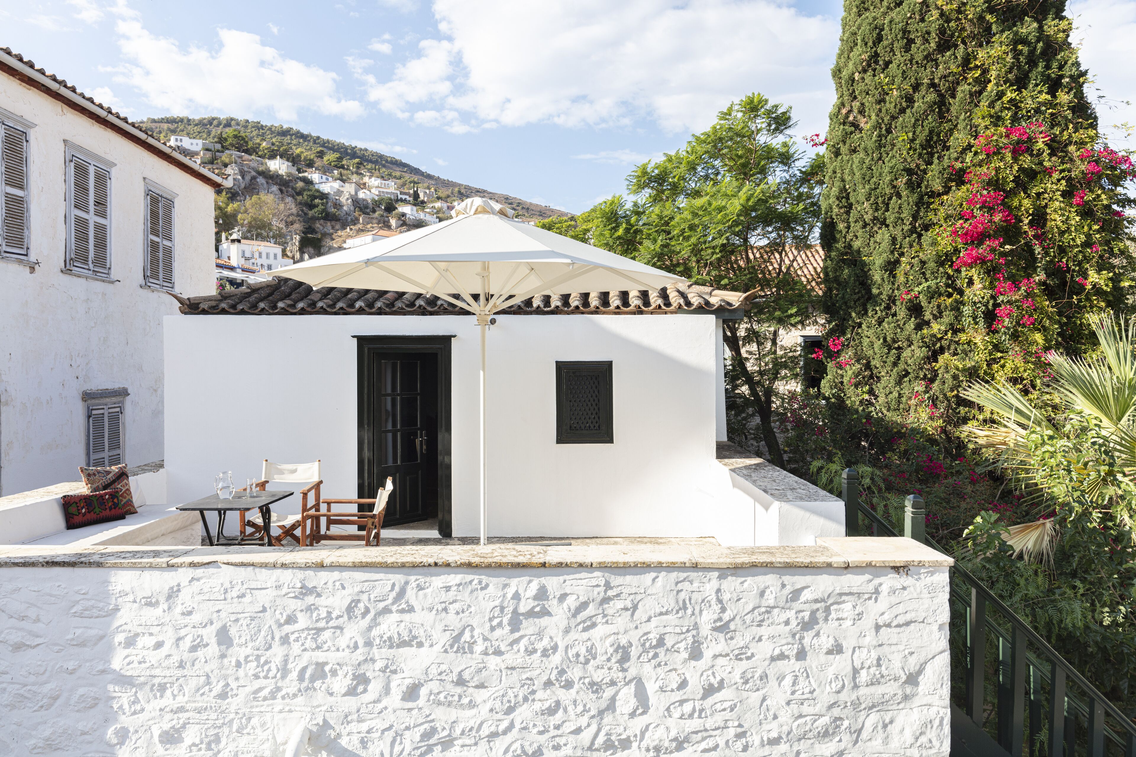 Um edifício branco com telhado de azulejos, um pequeno pátio com mesa e cadeiras e uma parede de pedra.