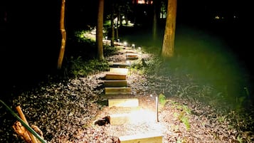 Uma trilha com degraus serpenteia por uma área arborizada, iluminada por luzes.