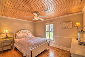 Quarto com teto de madeira, cama com cabeceira branca, mesa de cabeceira com abajur, janela com persianas e um objeto decorativo na parede.