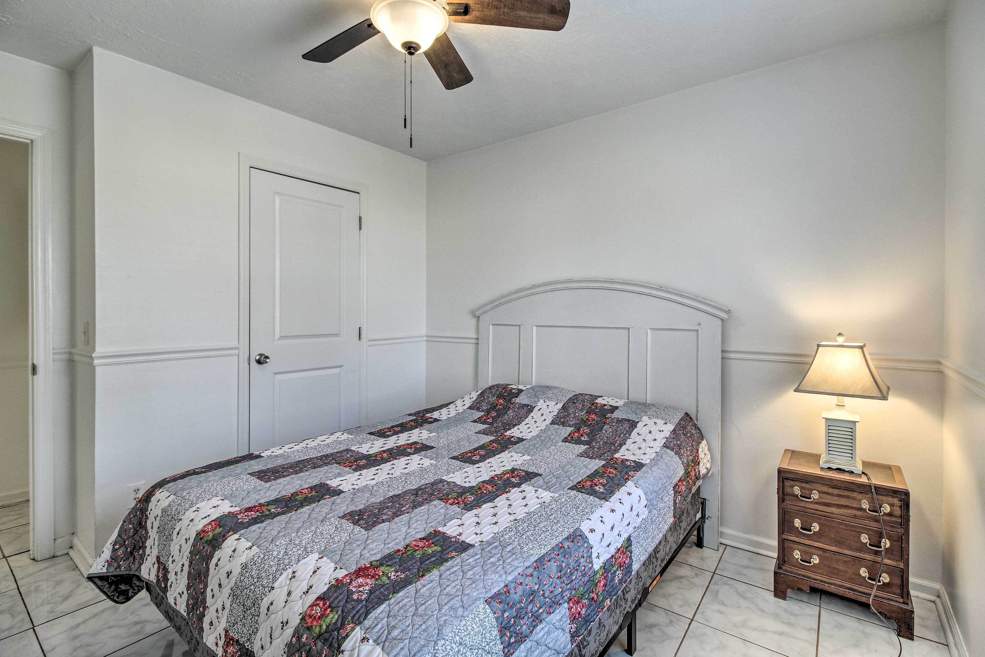 Quarto com cama, mesa de cabeceira, abajur, ventilador de teto e porta.