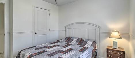 Quarto com cama, mesa de cabeceira, abajur, ventilador de teto e porta.