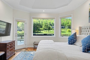 Quarto com uma cama grande, duas janelas com vista para uma paisagem verde, uma cômoda e uma televisão.
