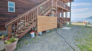 Uma cabana de madeira com uma escadaria também de madeira que dá para um deck, localizada em uma área de cascalho com vista para a água e montanhas.