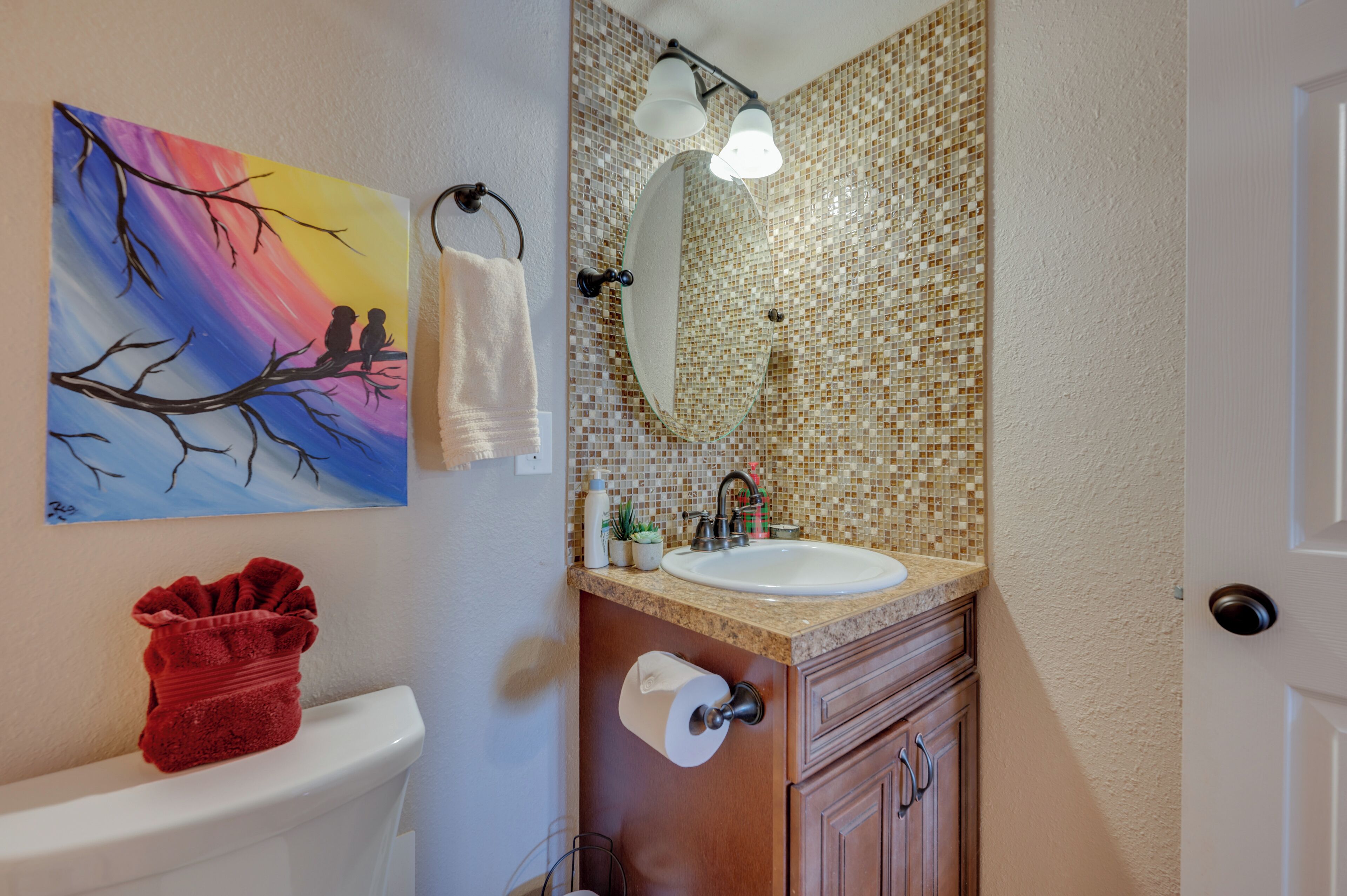 Casa de banho com painel de azulejos, uma pia, espelho, sanita e um quadro com pássaros num ramo.